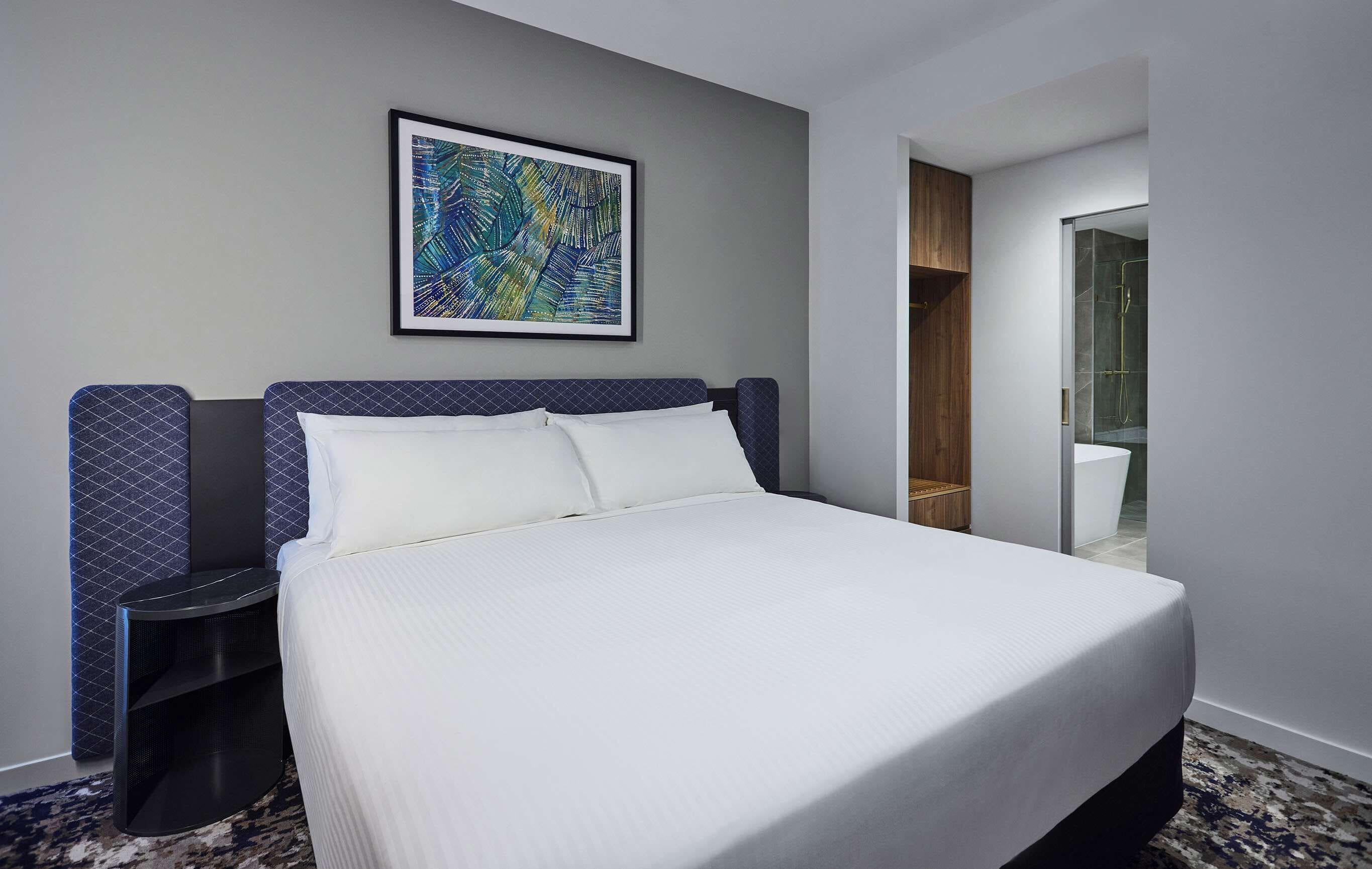 Quarto de hotel moderno com uma cama grande, uma pintura abstrata emoldurada, um espaldar azul escuro e um banheiro visível.