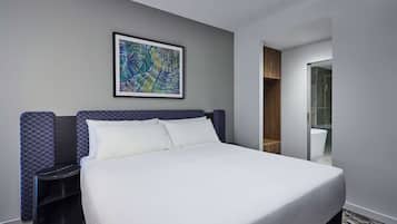 Quarto de hotel moderno com uma cama grande, uma pintura abstrata emoldurada, um espaldar azul escuro e um banheiro visível.