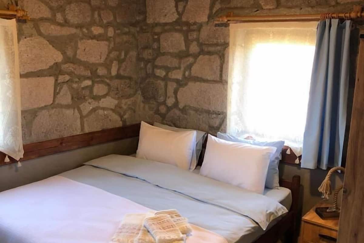 Quarto com parede de pedra, cama com roupa de cama branca, cabeceira de madeira, mesa de cabeceira com luminária e janela com cortinas.