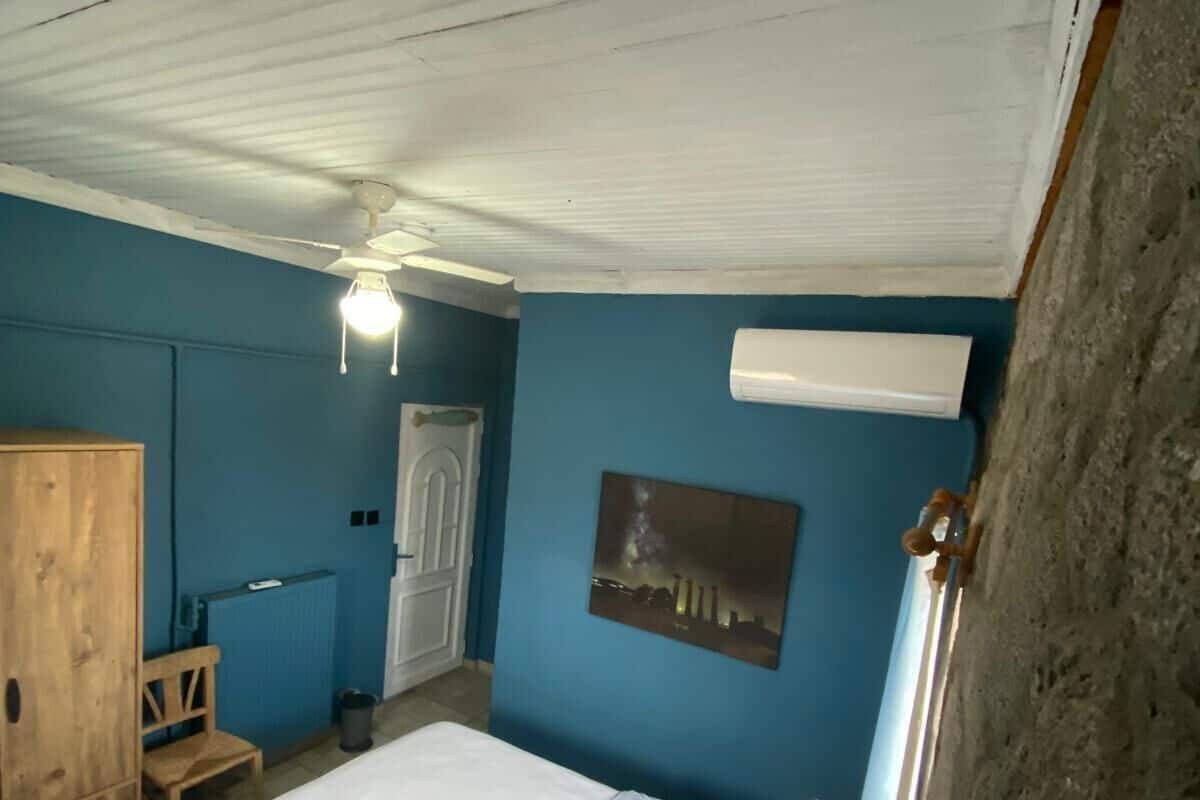 Quarto com cama, guarda-roupa de madeira, radiador, porta branca e ventoinha no teto.