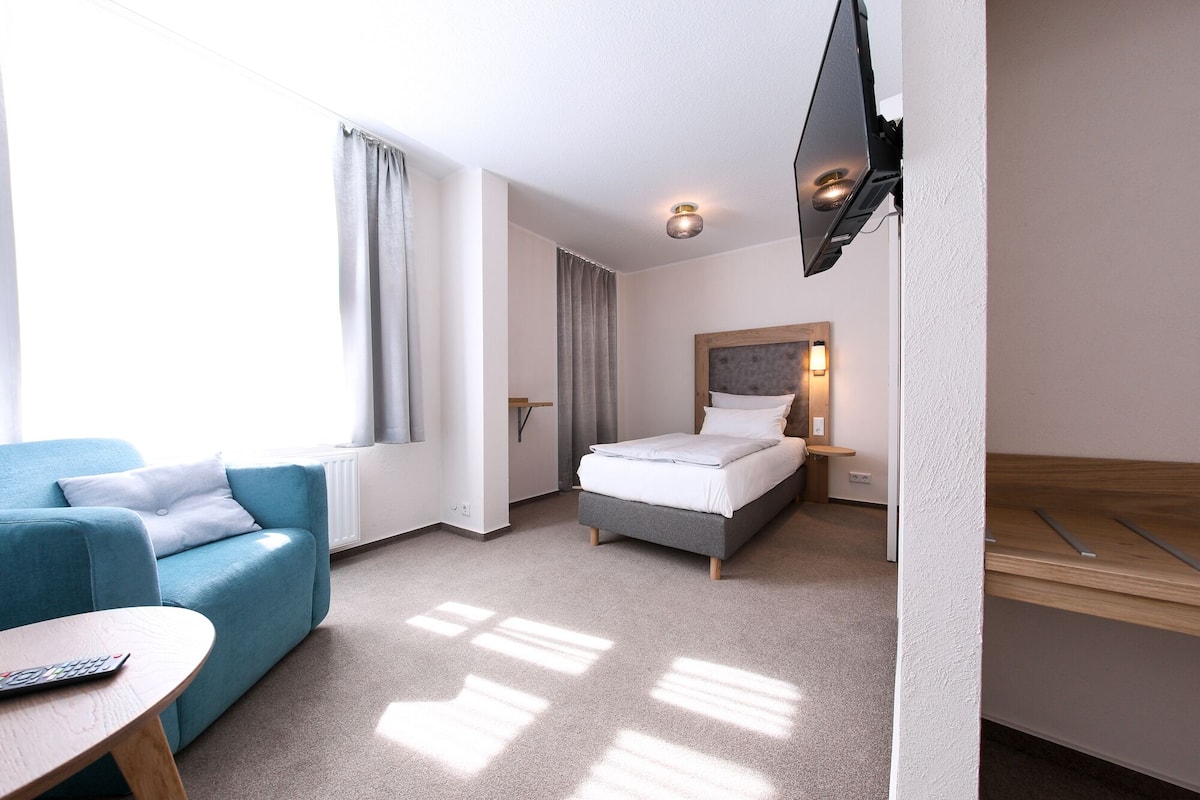 Quarto de hotel com uma cama, um sofá azul, uma mesinha e uma TV fixada na parede.