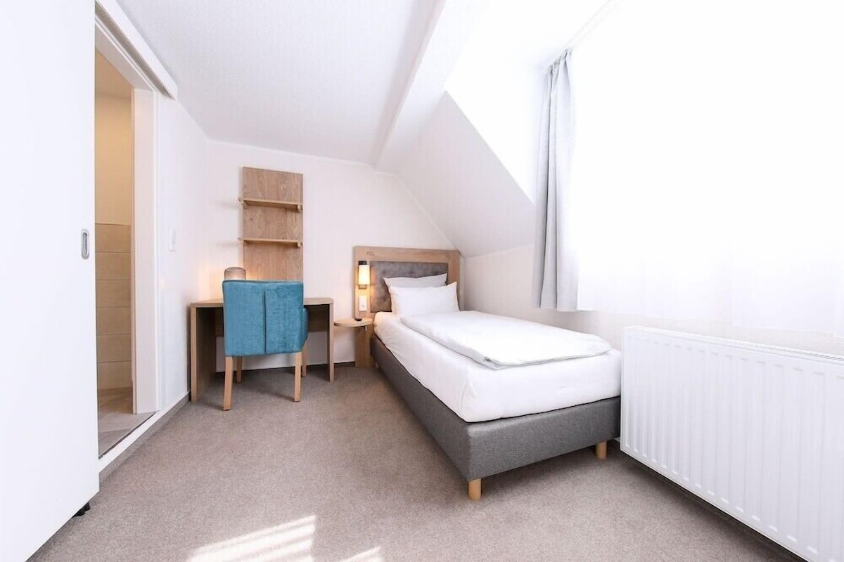 Quarto pequeno e bem iluminado, com uma cama, uma escrivaninha e uma cadeira. Há uma janela que proporciona luz natural, e um radiador é visível na parede.