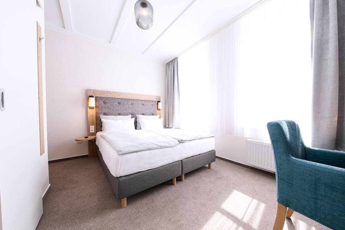 Quarto de hotel moderno com uma cama grande, uma mesa de cabeceira, uma cadeira e uma janela com cortinas.