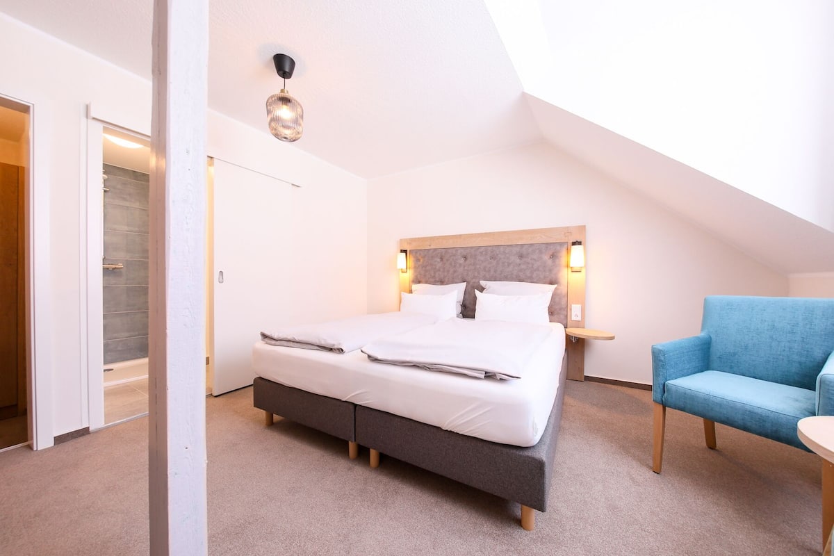 Quarto de hotel moderno com uma cama grande, uma poltrona azul e um claraboia.