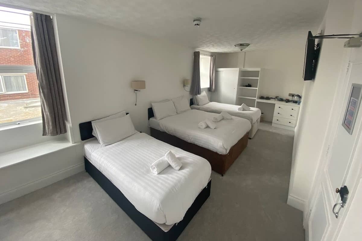 Quarto de hotel com três camas, uma mesa, um guarda-roupa e uma janela com cortinas.