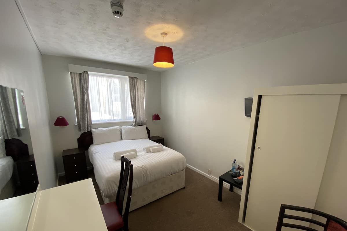 Quarto de hotel com cama, escrivaninha, cadeira, janela com cortinas, luminária pendente vermelha e porta.