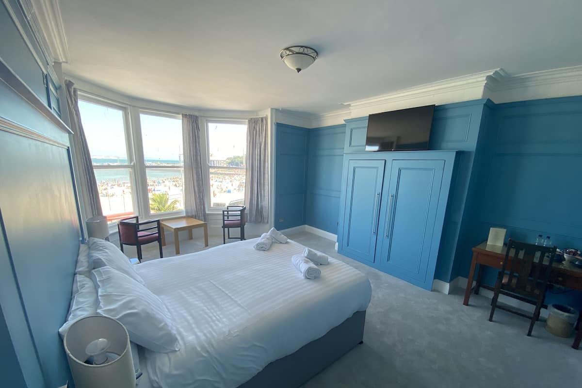 Quarto de hotel com uma cama grande, uma escrivaninha, uma cadeira e uma televisão. Há uma janela com vista para a praia.