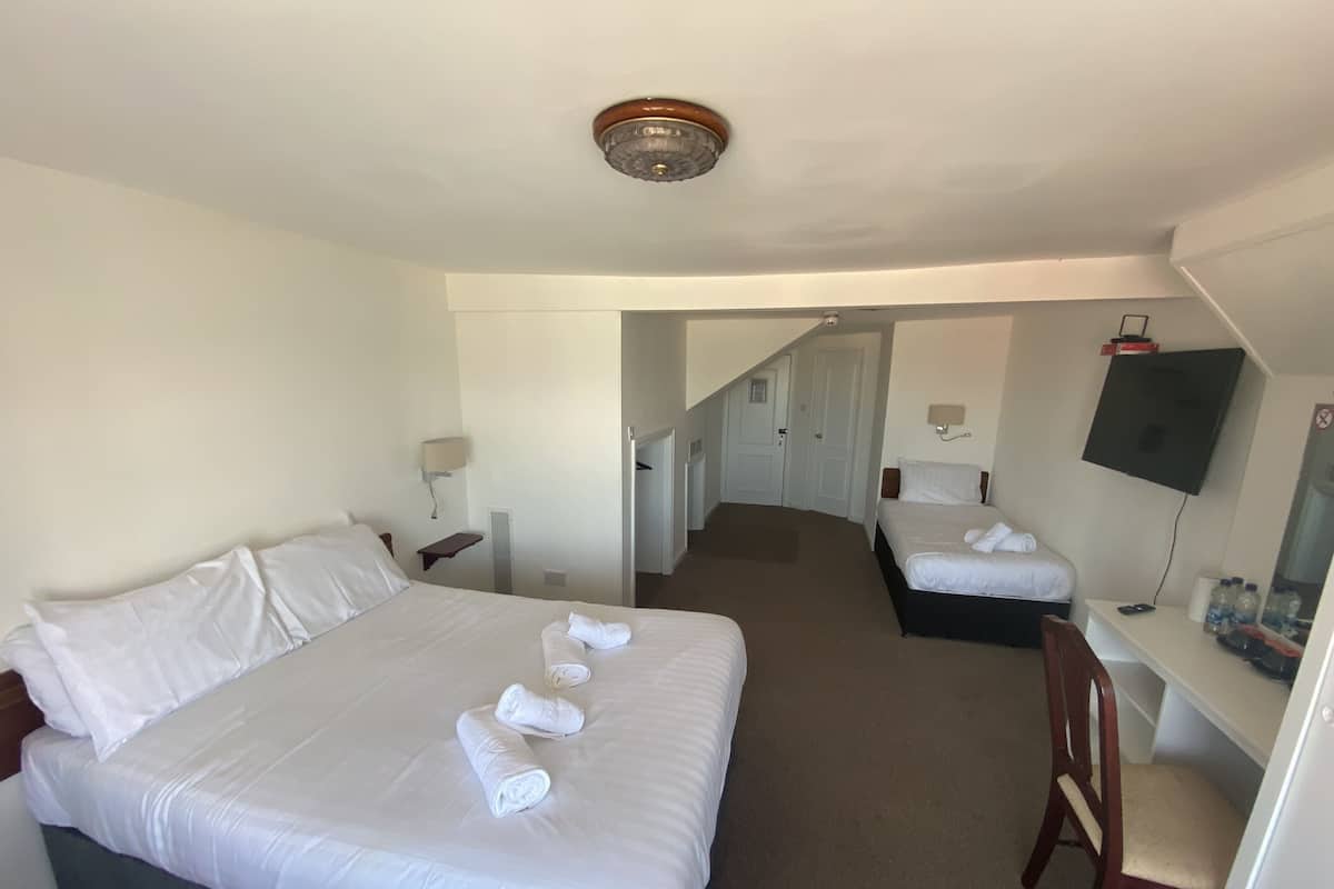 Quarto de hotel com uma cama grande, uma cama menor, uma escrivaninha com cadeira, televisão e um banheiro visível por uma porta aberta.