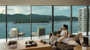 Sala de estar moderna com uma grande janela com vista para um corpo de água e uma colina arborizada. Há um sofá, uma mesa de centro e uma mesinha com uma planta.