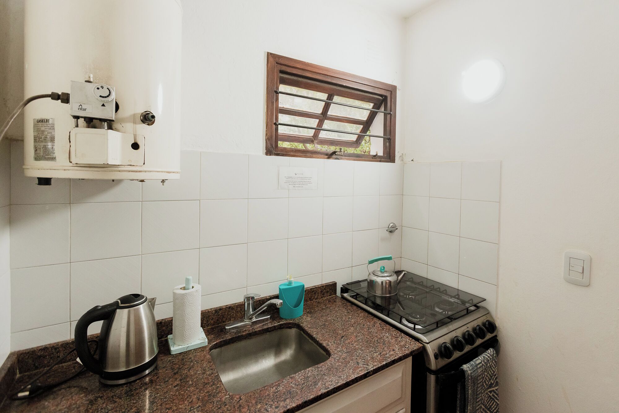 Uma cozinha com azulejos brancos, fogão a gás, chaleira, pia e janela com moldura de madeira.