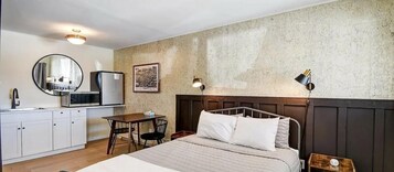 Quarto de hotel com cama, mesa de cabeceira, luminária, escrivaninha com cadeira e área de cozinha compacta.