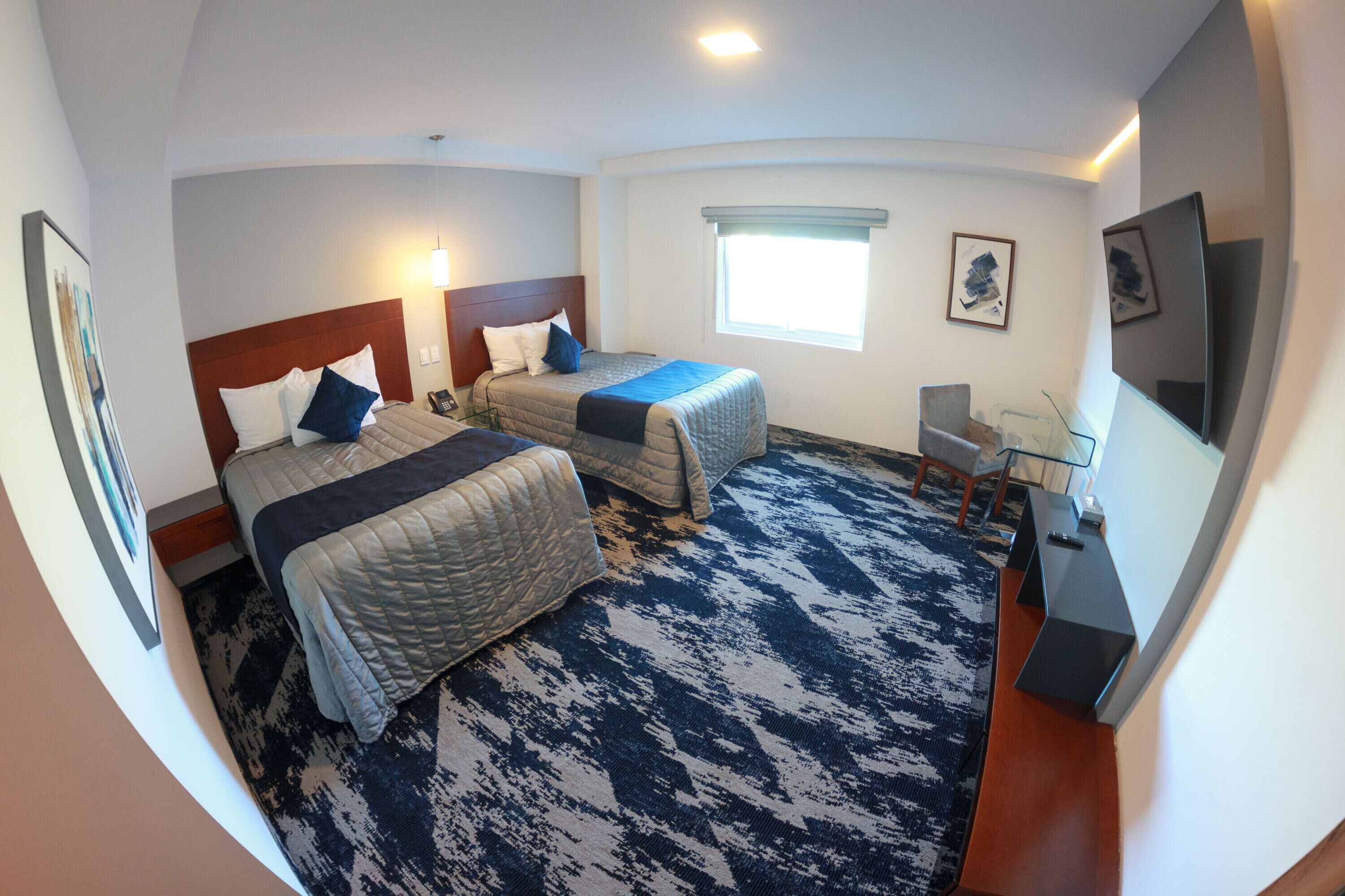 Quarto de hotel com duas camas, um tapete grande, uma escrivaninha, uma cadeira e uma televisão.