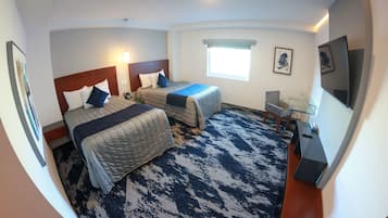 Quarto de hotel com duas camas, um tapete grande, uma escrivaninha, uma cadeira e uma televisão.