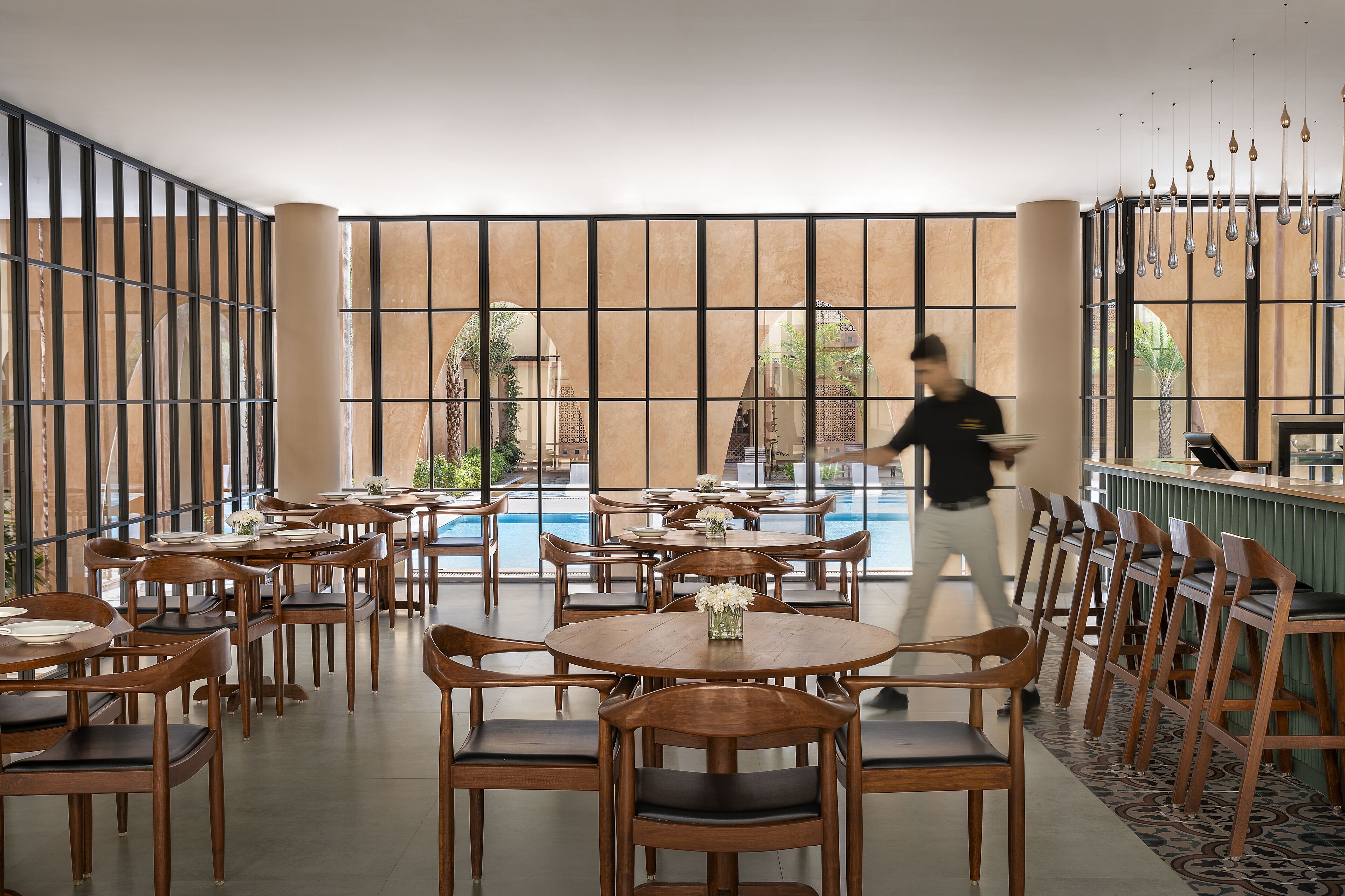 Uma ampla sala de jantar com várias mesas redondas e cadeiras de madeira, grandes janelas com vista para a piscina e área externa, e um balcão de bar com banquetas.