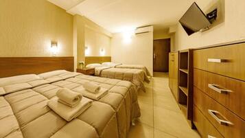 Quarto de hotel com três camas, televisão fixada na parede e guarda-roupa de madeira.