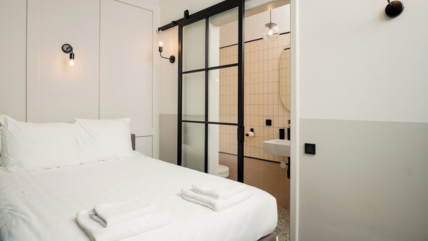 Quarto de hotel com cama, porta deslizante para o banheiro e luminárias fixadas na parede.