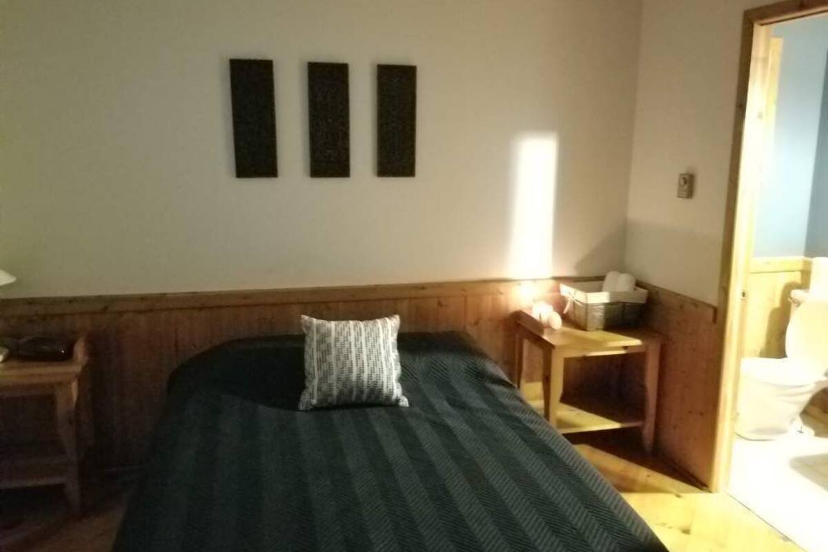 Quarto com cama, cabeceira de madeira, mesa de cabeceira, pequena mesa e prateleira fixada na parede.