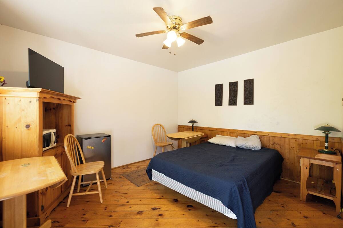 Quarto com cama, escrivaninha, cadeira, mesinha, ventoinha no teto e micro-ondas.