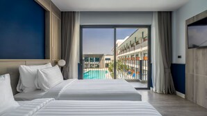 Quarto de hotel com duas camas, uma janela ampla com vista para uma piscina e edifícios, e uma televisão de tela plana presa à parede.