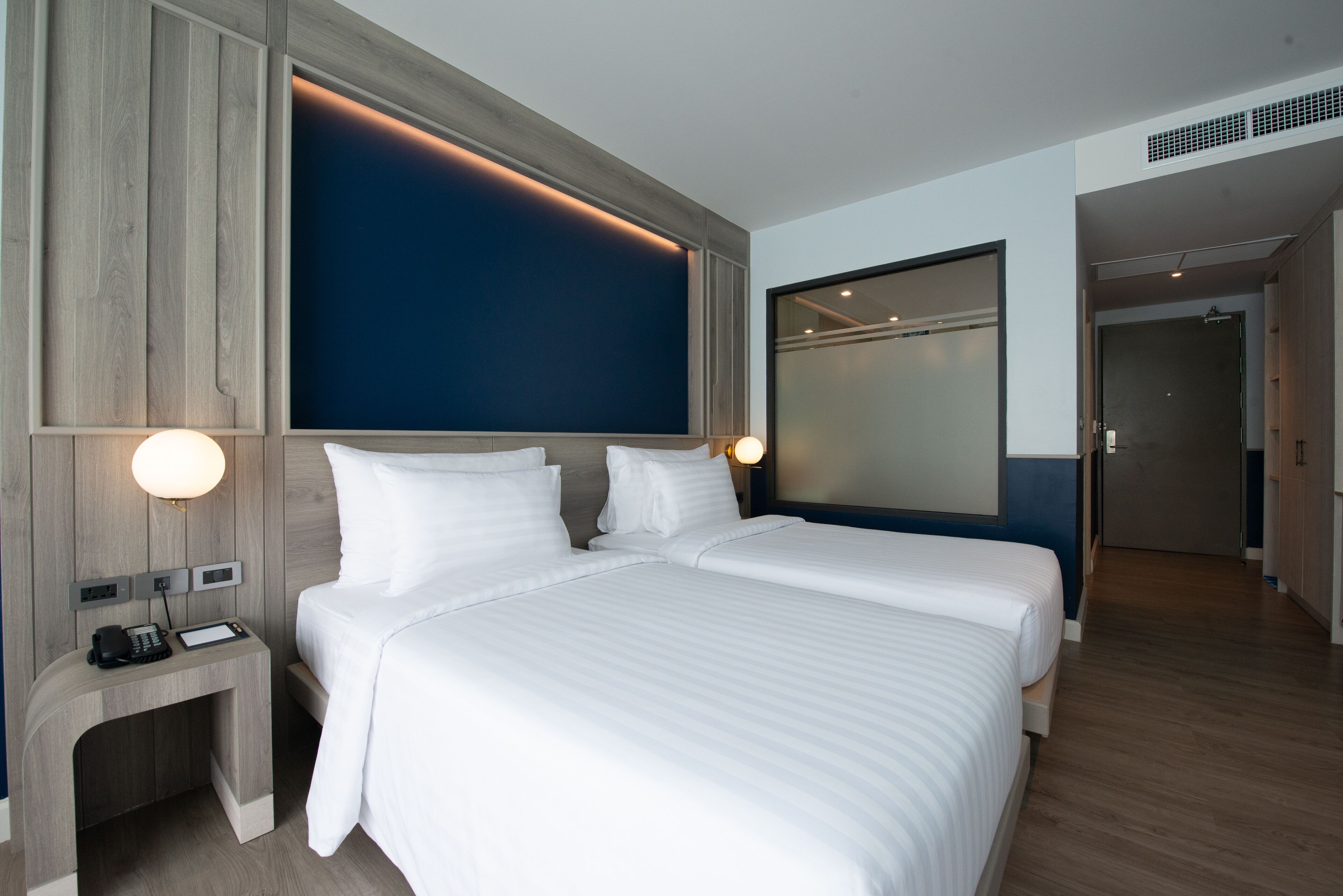 Quarto de hotel com duas camas, uma cabeceira de madeira, mesa de cabeceira com telefone e luminária, e uma janela grande com vista.