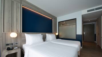 Quarto de hotel com duas camas, uma cabeceira de madeira, mesa de cabeceira com telefone e luminária, e uma janela grande com vista.