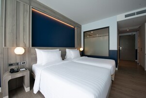 Quarto de hotel com duas camas, uma cabeceira de madeira, mesa de cabeceira com telefone e luminária, e uma janela grande com vista.