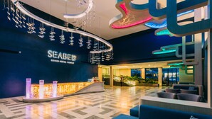 Um saguão de hotel com balcão de recepção, área de estar e escadaria que leva aos andares superiores.