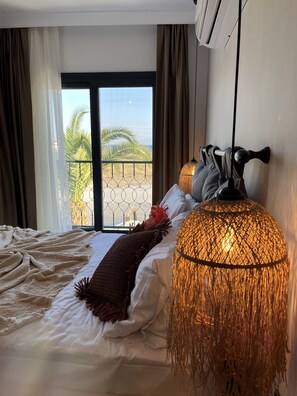 Quarto com uma cama grande, luminária pendente e vista para palmeiras pela janela.