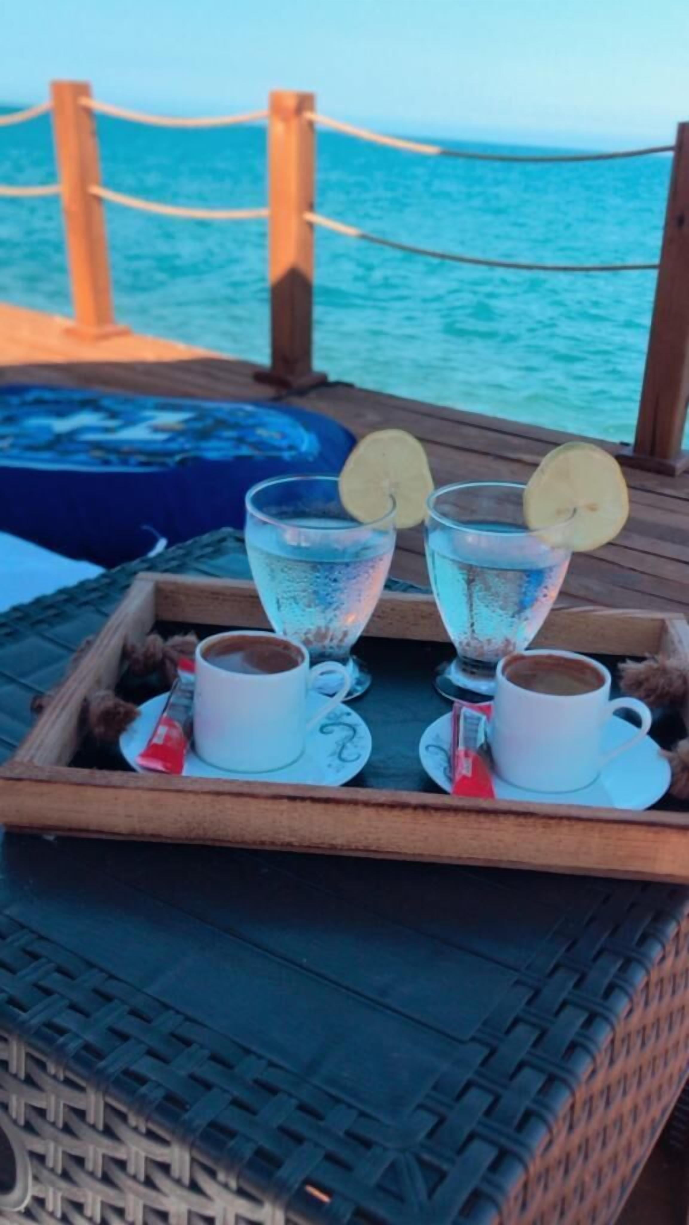 Uma bandeja com duas xícaras de café e dois copos de água sobre um deck de madeira com vista para o oceano.