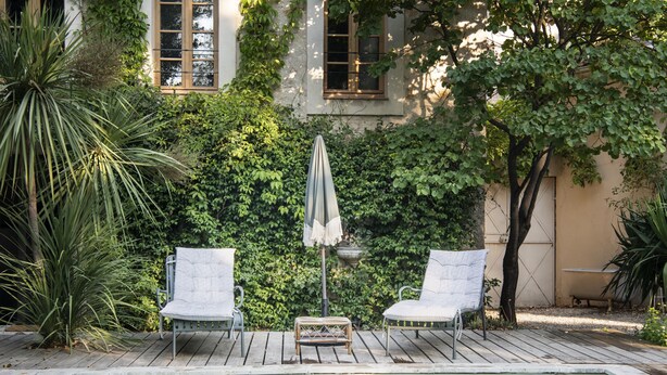 Área de piscina com duas cadeiras e um guarda-sol, cercada por vegetação exuberante e um edifício com janelas.