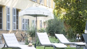 Um jardim com três cadeiras de descanso e uma mesa, um guarda-sol branco, e um edifício com persianas ao fundo.