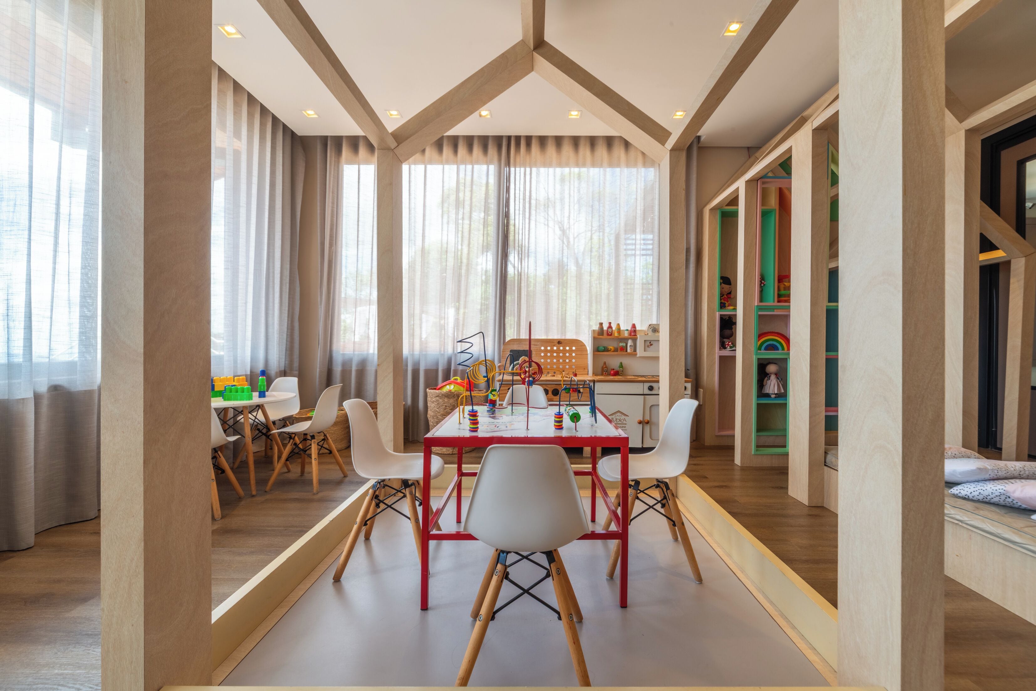 Uma área de jantar moderna com mesa vermelha e cadeiras brancas, uma cozinha ao fundo e um espaço infantil com prateleiras coloridas.
