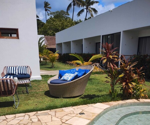Uma área à beira da piscina com mobiliário de vime, uma piscina pequena e um edifício com fachada branca e colunas.
