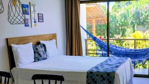 Quarto de hotel com cama, uma mesa de jantar com prato e xícara, rede do lado de fora da janela e vista para área verde.