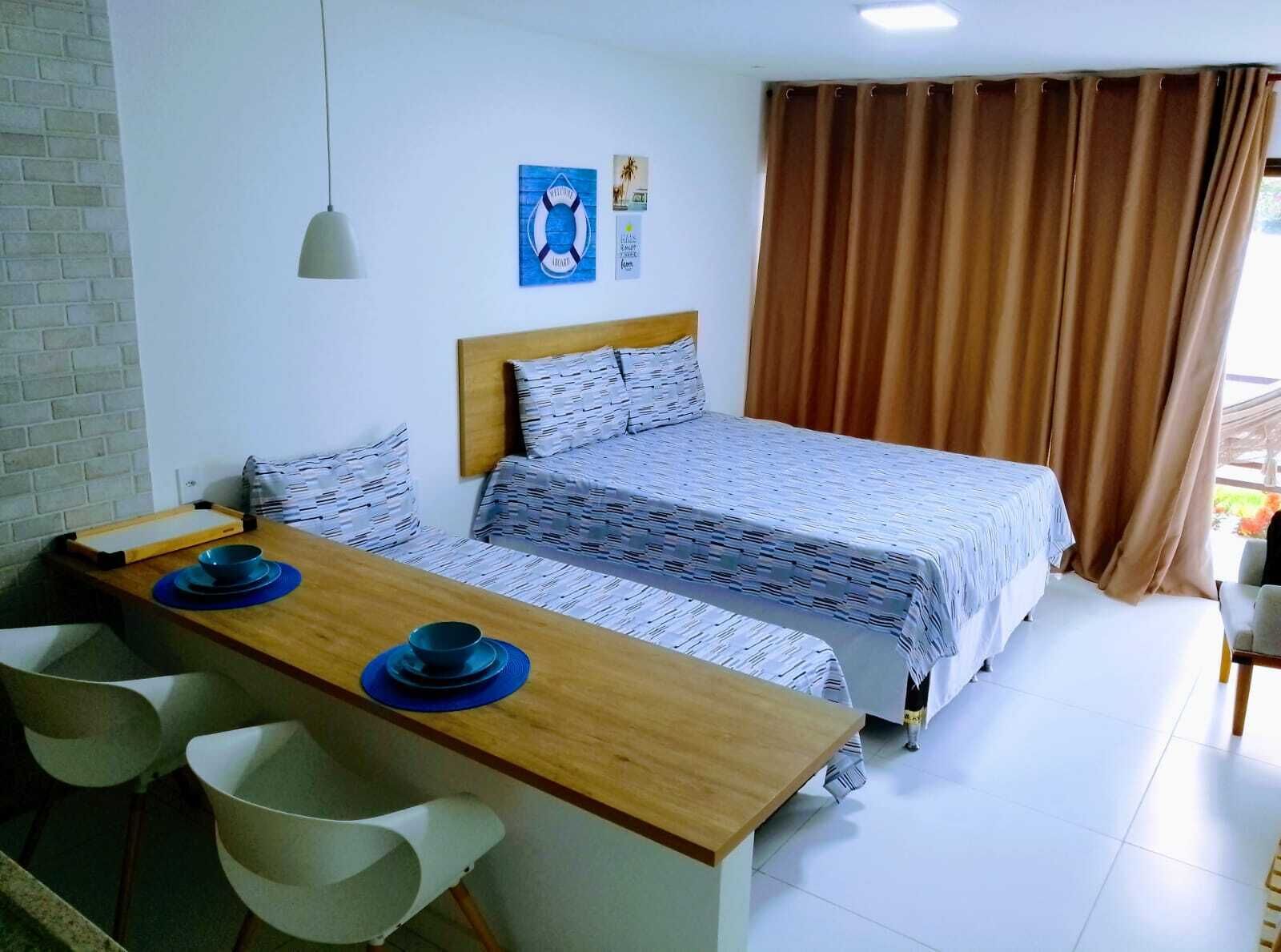Quarto de hotel com cama, escrivaninha de madeira com cadeira, mesinha com uma tigela e um quadro decorativo de boia.