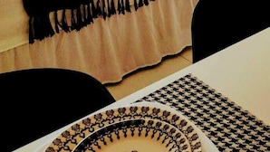 Um quarto com cama, mesa com pratos, cadeira, janela com cortinas e um banquinho.