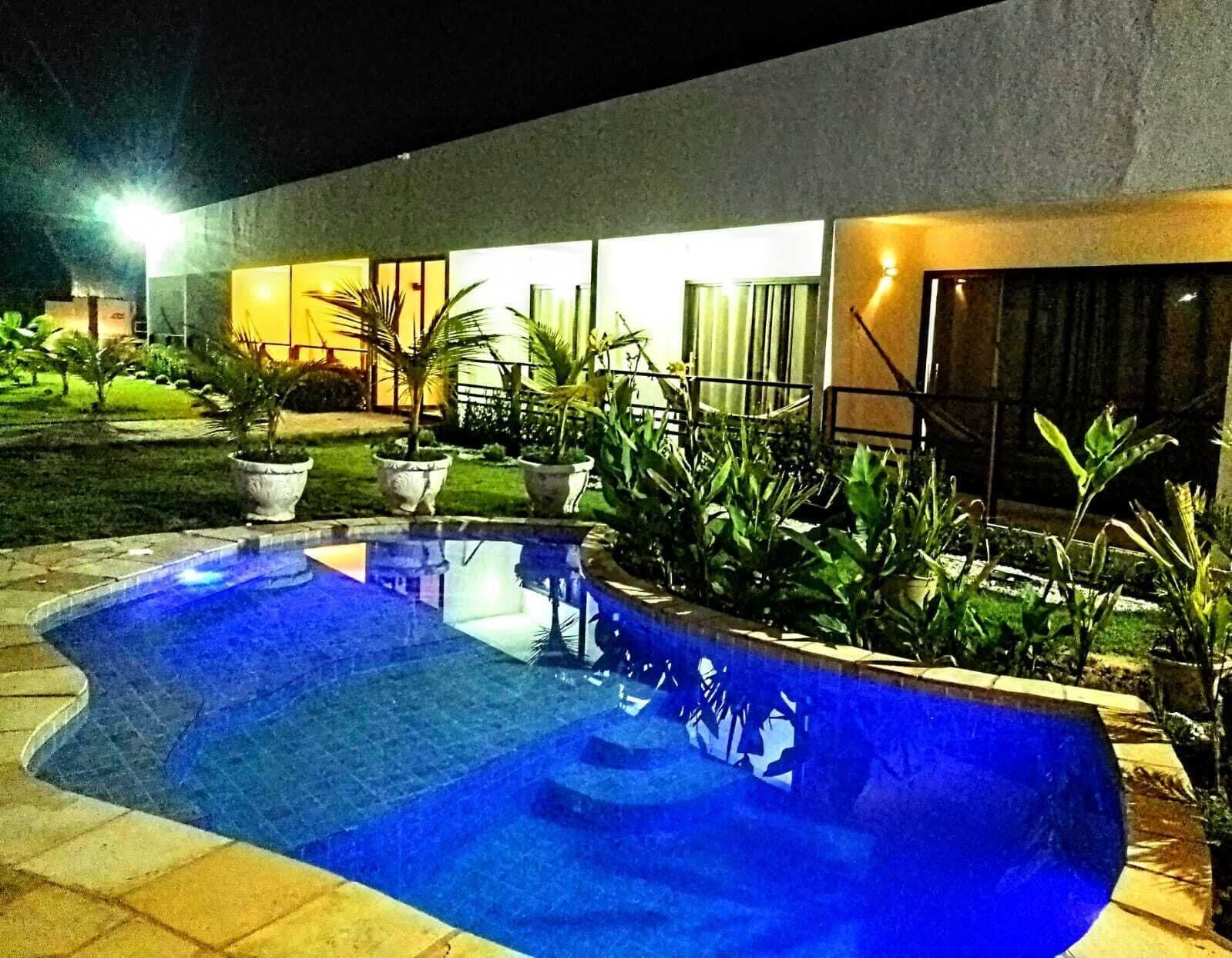Piscina com deck ladrilhado, rodeada por vasos de plantas e um edifício bem iluminado ao fundo.