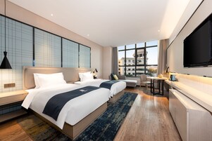 Quarto de hotel com duas camas, uma escrivaninha, uma cadeira, uma televisão e uma janela grande.