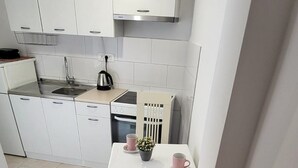 Uma cozinha pequena com armários brancos, um fogão e uma mesa com duas chávenas cor-de-rosa e uma pequena planta.