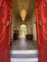 Uma entrada imponente com portas vermelhas, um candelabro e vista de um interior espaçoso com uma escadaria para uma área iluminada.