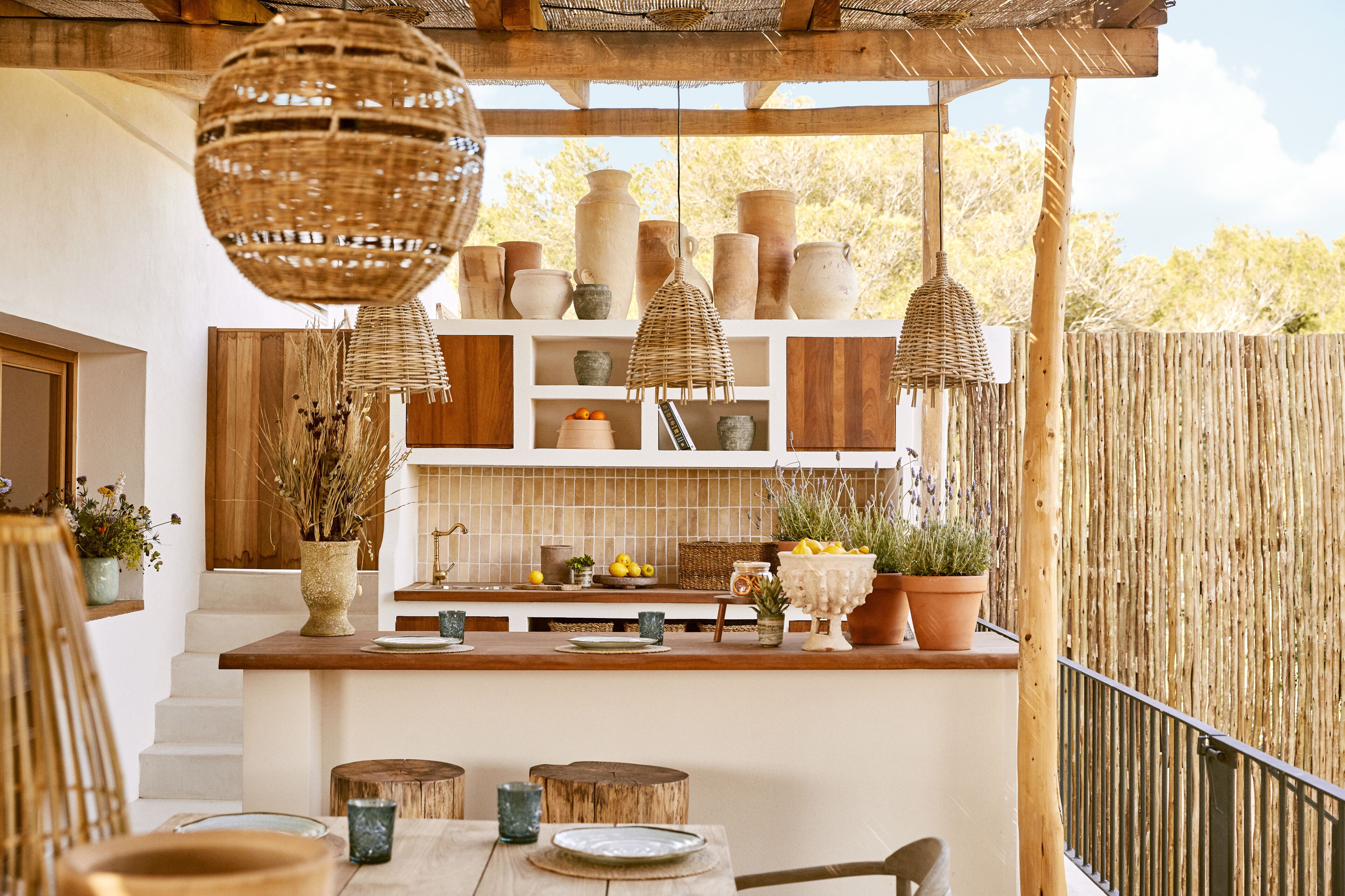 Uma cozinha rústica ao ar livre, com prateleiras de madeira, luminárias de vime suspensas e plantas em vasos.