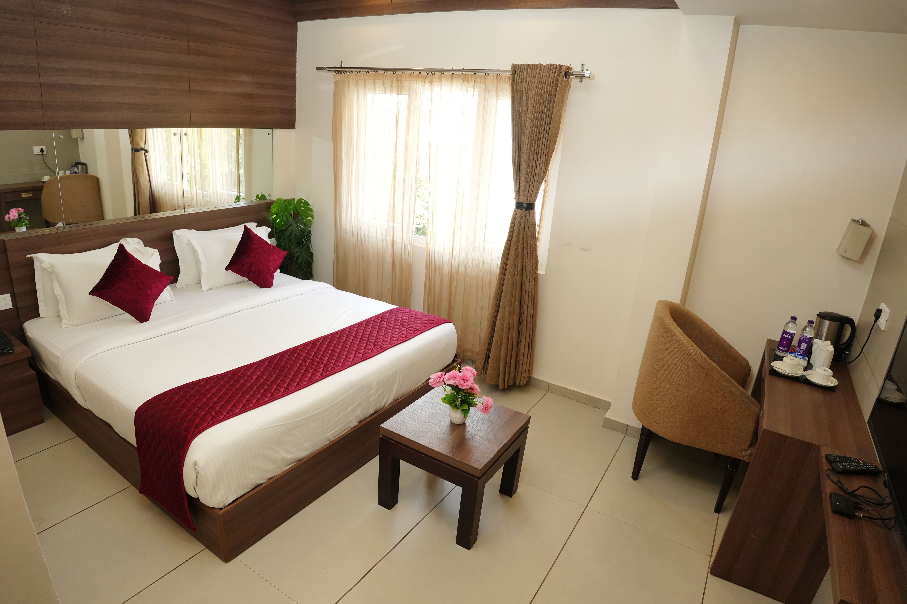 Quarto de hotel com uma cama grande, uma mesa pequena com flores, uma cadeira e uma escrivaninha com cafeteira e garrafas.