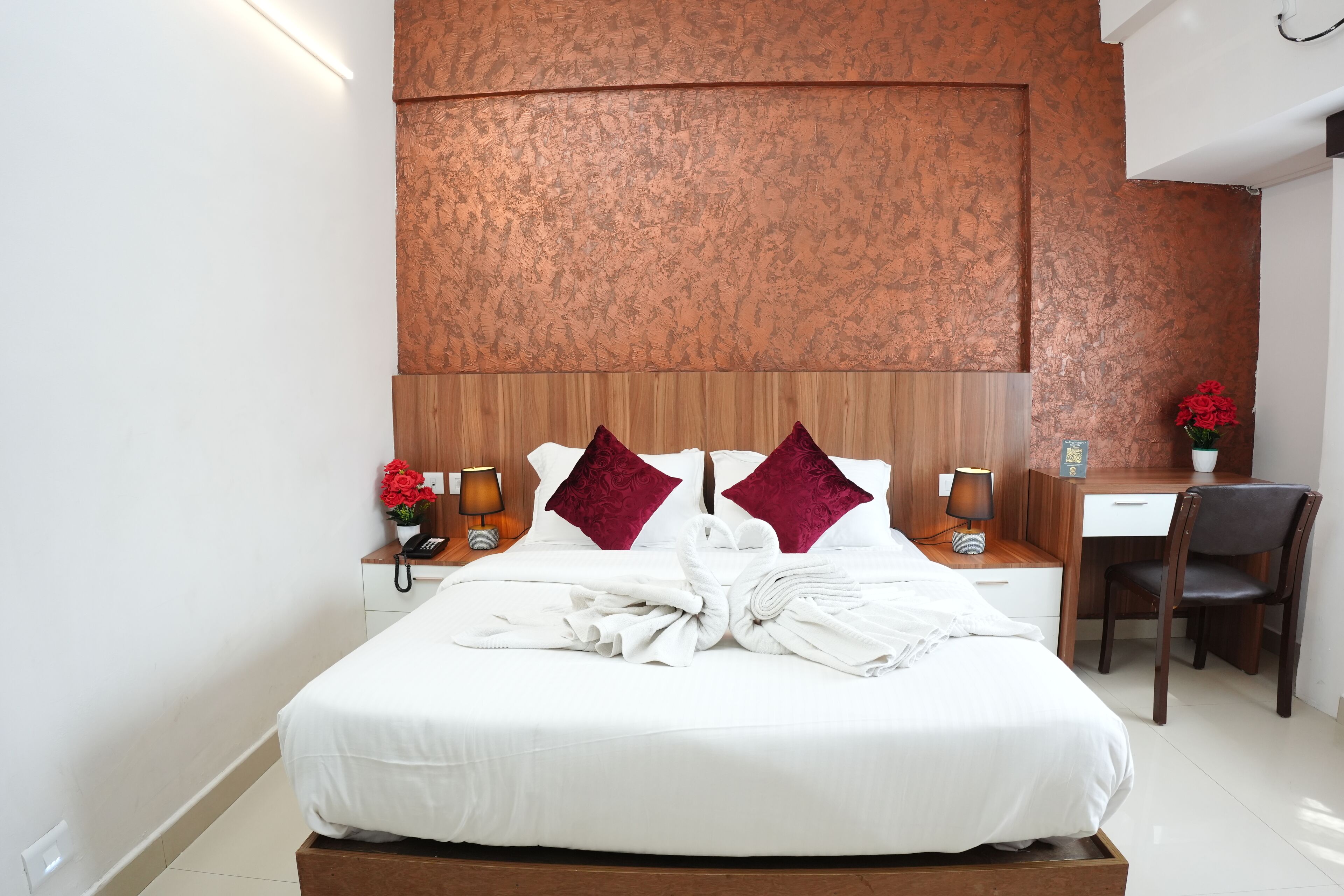 Quarto de hotel com cama bem arrumada, cabeceira de madeira, escrivaninha com cadeira e mesa de cabeceira com abajur e telefone.