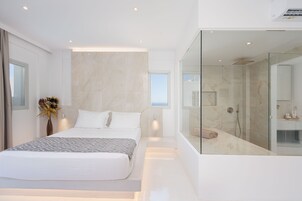 Quarto de hotel moderno com uma cama grande, box de vidro para o chuveiro e vista do céu.
