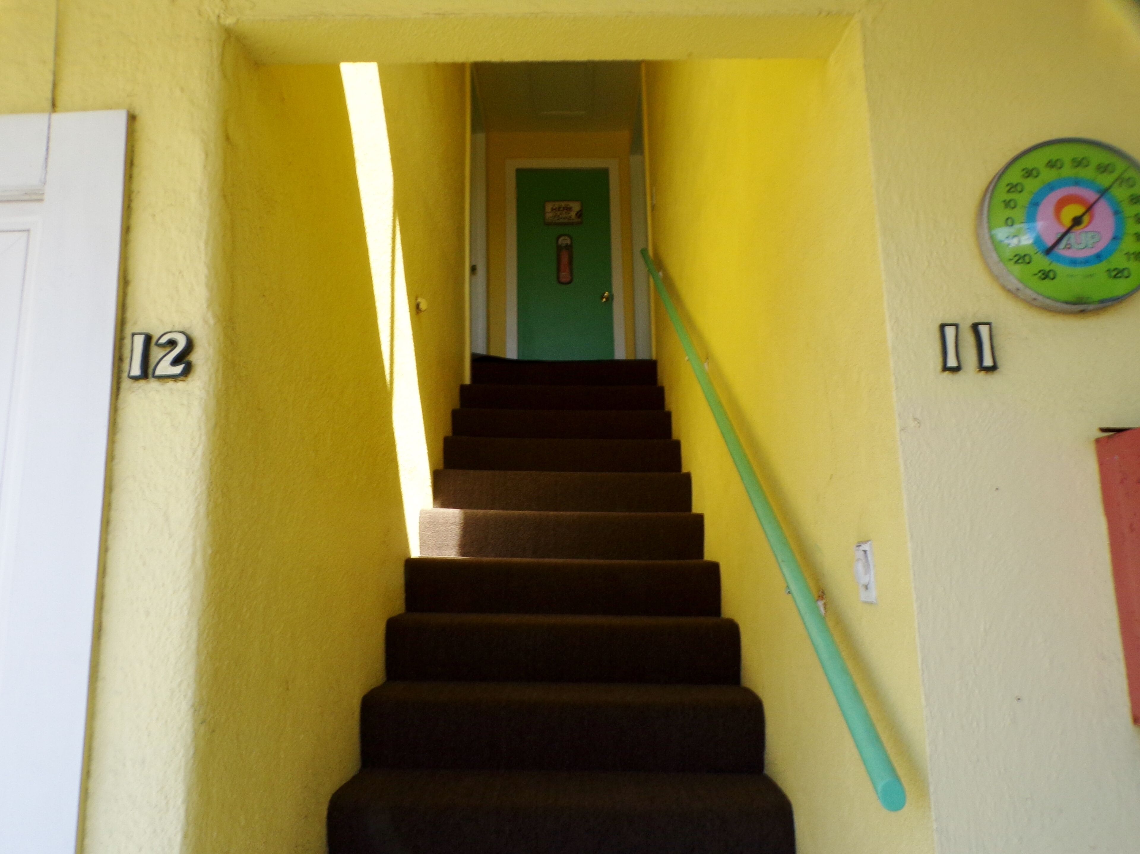 Uma escada leva a uma porta com uma placa verde, os números 12 e 11 nas paredes, e um relógio à direita.