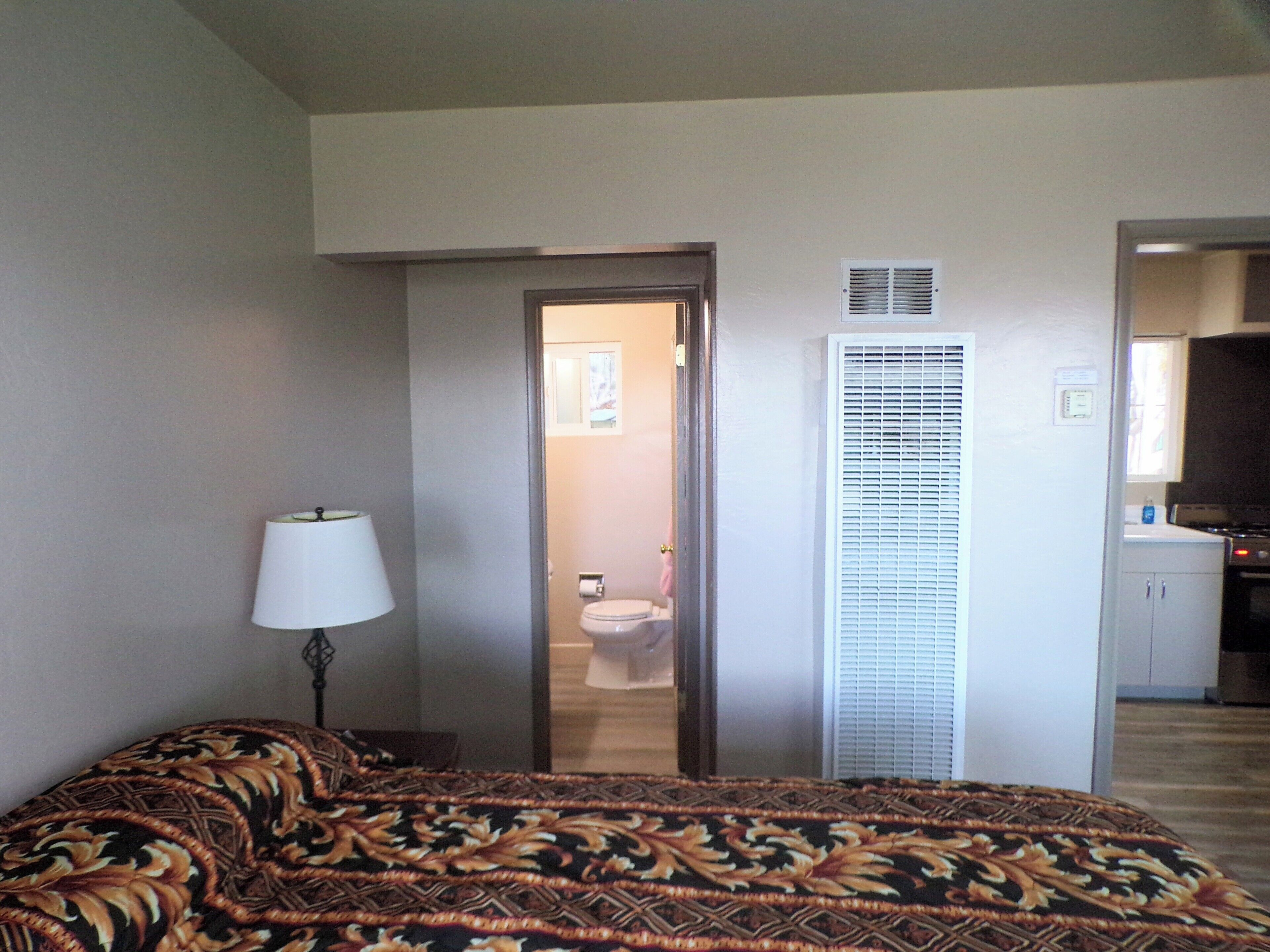 Quarto de hotel com cama, luminária, porta para banheiro e cozinha compacta.
