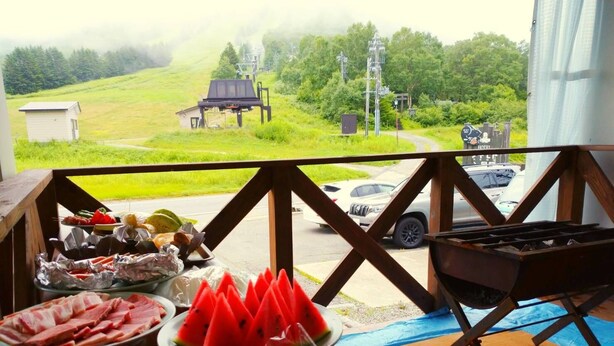 Varanda com churrasqueira, diversos alimentos e vista para paisagem rural com um hotel e carros estacionados.