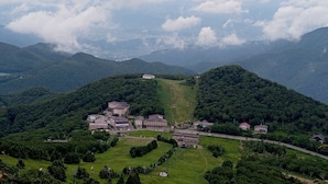 Uma área montanhosa com um conjunto de edifícios, provavelmente um resort ou hotel, cercada por vegetação exuberante e uma estrada que leva até ele.