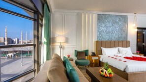 Quarto de hotel com uma cama grande, um sofá, uma cadeira, uma pequena mesa com frutas e vista para um horizonte urbano com edifícios e uma ponte.