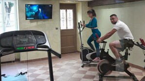 Sala de ginástica com bicicletas ergométricas, esteira e televisão exibindo um filme.